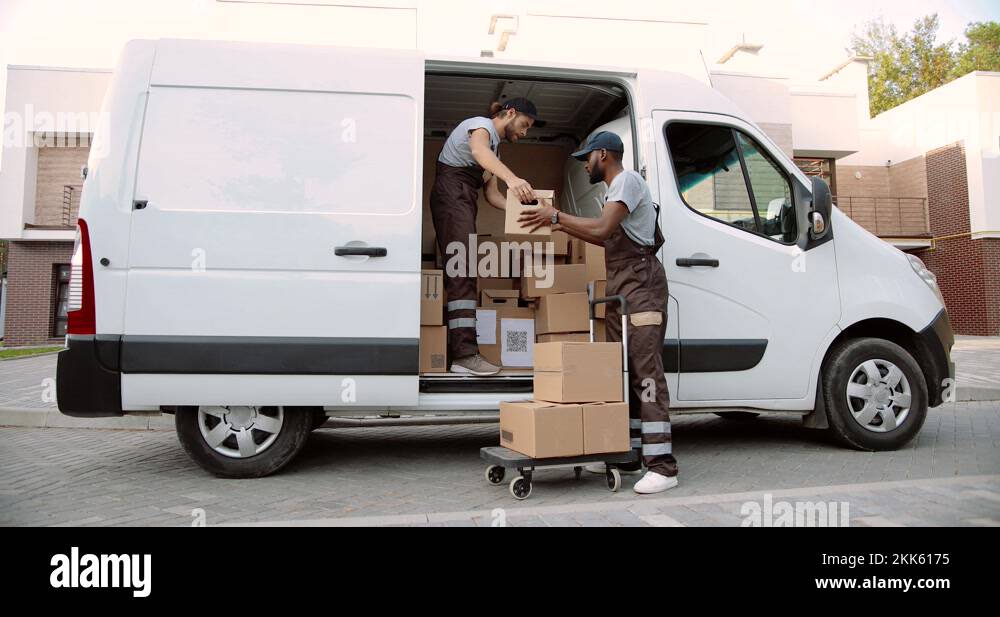 Young mixed-races delivery men coworkers taking boxes from van putting ...