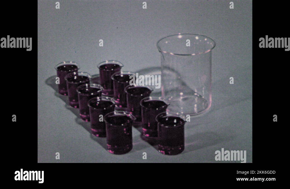 1950s: Ten small beakers of purple liquid sit next to empty beaker. Man ...