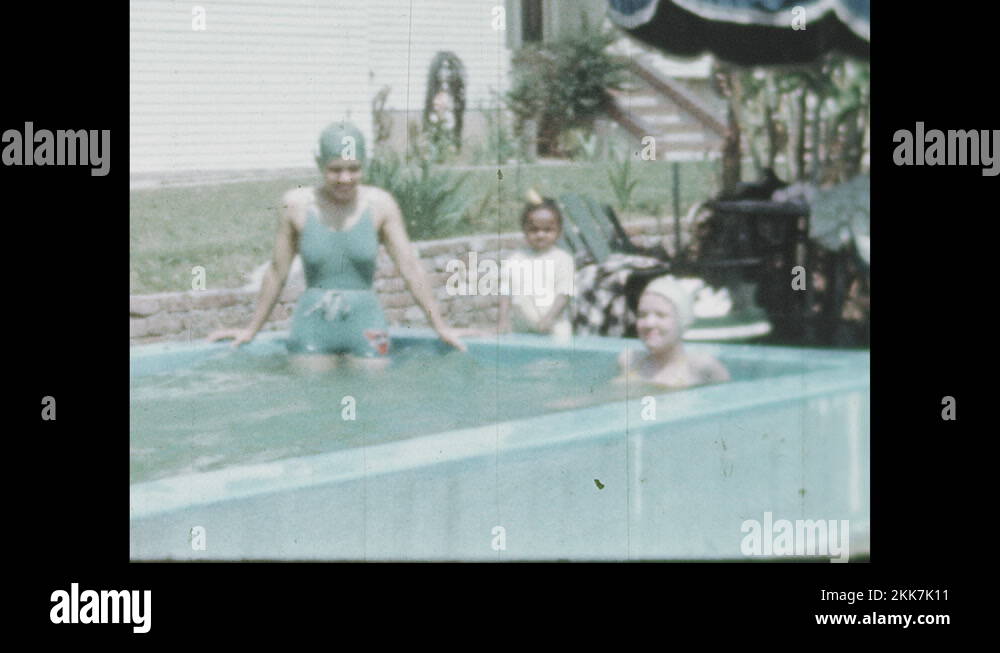 1950s: Two women swim in pool in yard of house. Man and woman sit on ...