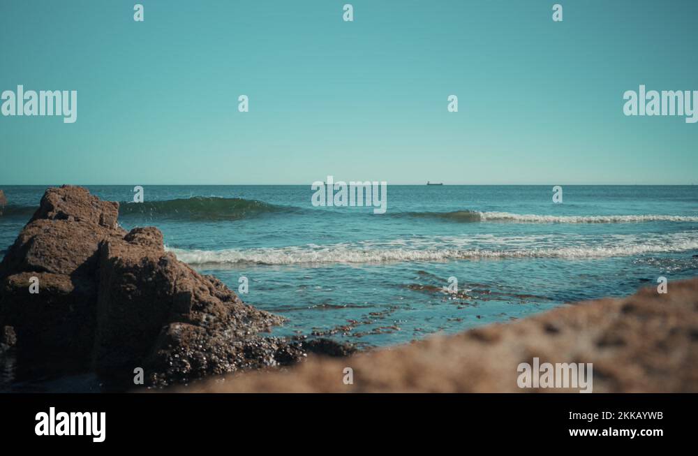 Ocean shoreline with cargo ships away and waves hit rock cliff gently ...