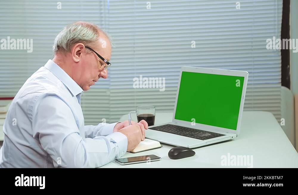 A man in glasses in an office with blinds at a computer works a green ...