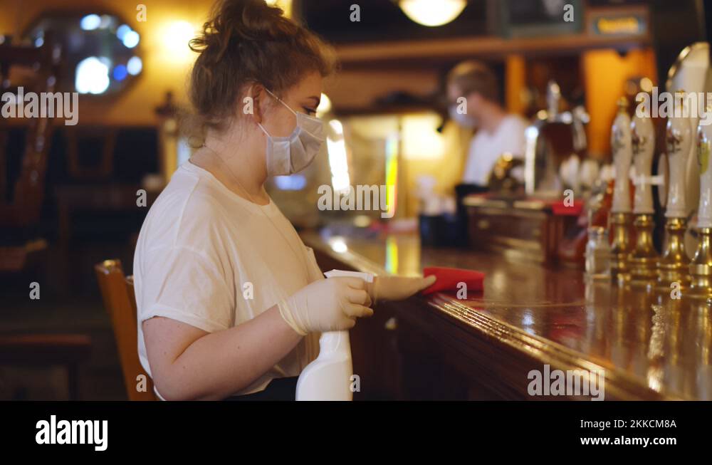 Young waitress in safety mask wiping bar counter with duster and ...