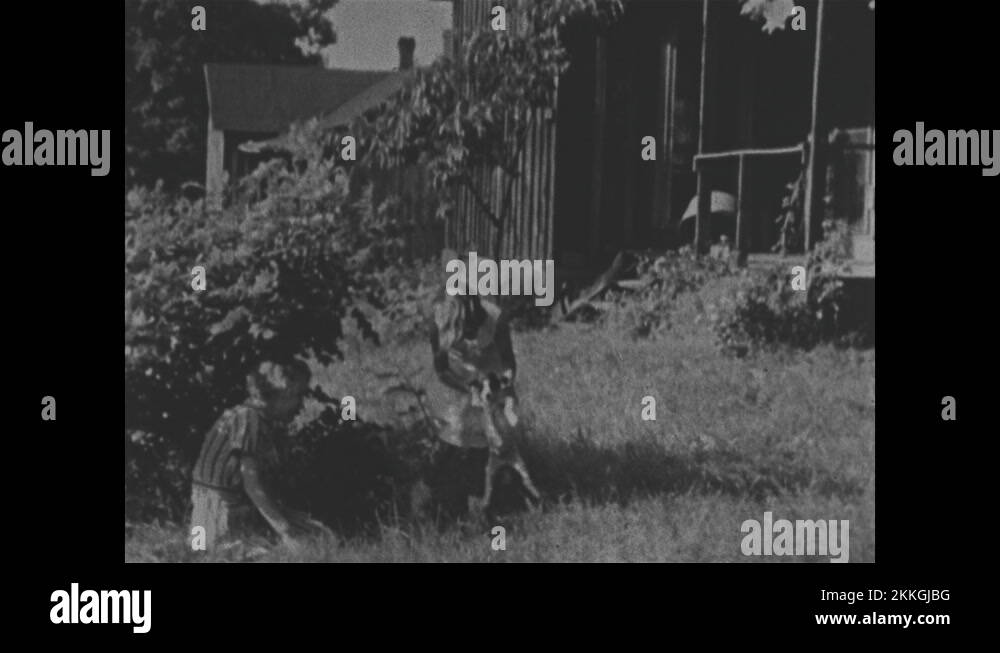 1940s: Children and woman play in sandbox. Girls sit under bush and ...
