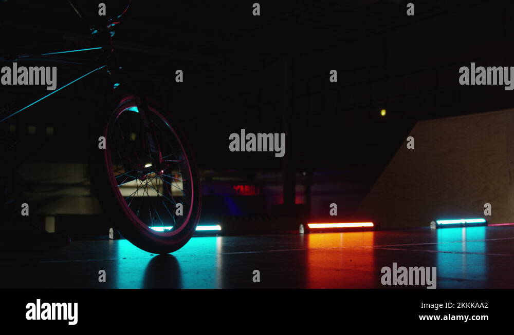 BMX rider walks bike across indoor ramps with neon lights on the ground ...