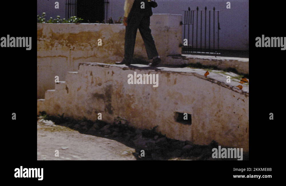 1970s: Man with camera walks up ramp and down steps. Archway, children ...