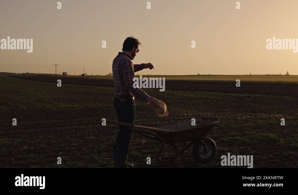 Male farmer making a rest of pushing wheelbarrow over farmland wearing ...