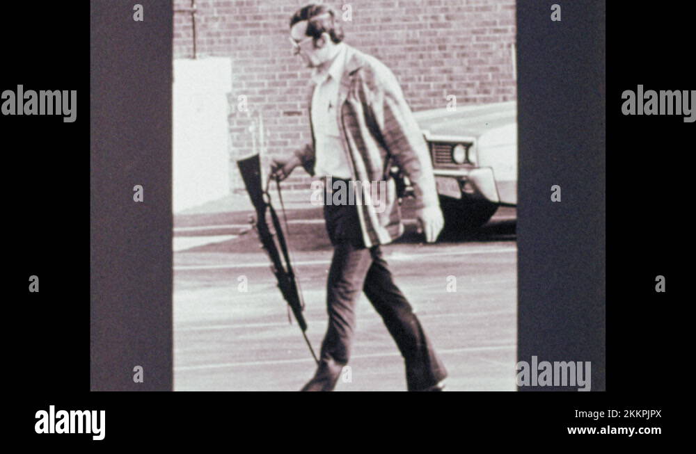 1970s: Still of man holding rifle by strap. Two cops hide behind cop ...