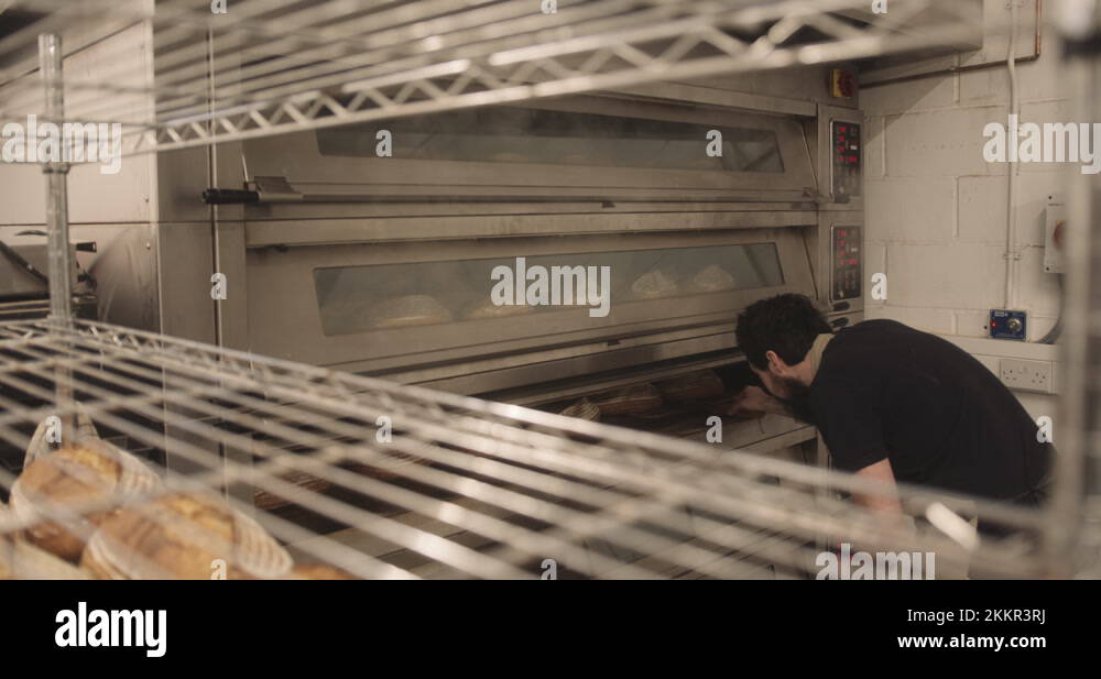 Bearded male baker taking baked sourdough bread out of the oven wearing ...