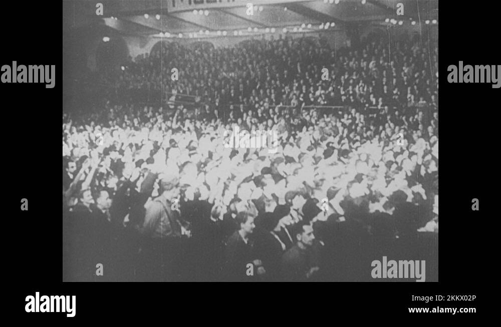 1930s Germany: Crowd of people cheer in auditorium. Adolf Hitler speaks ...