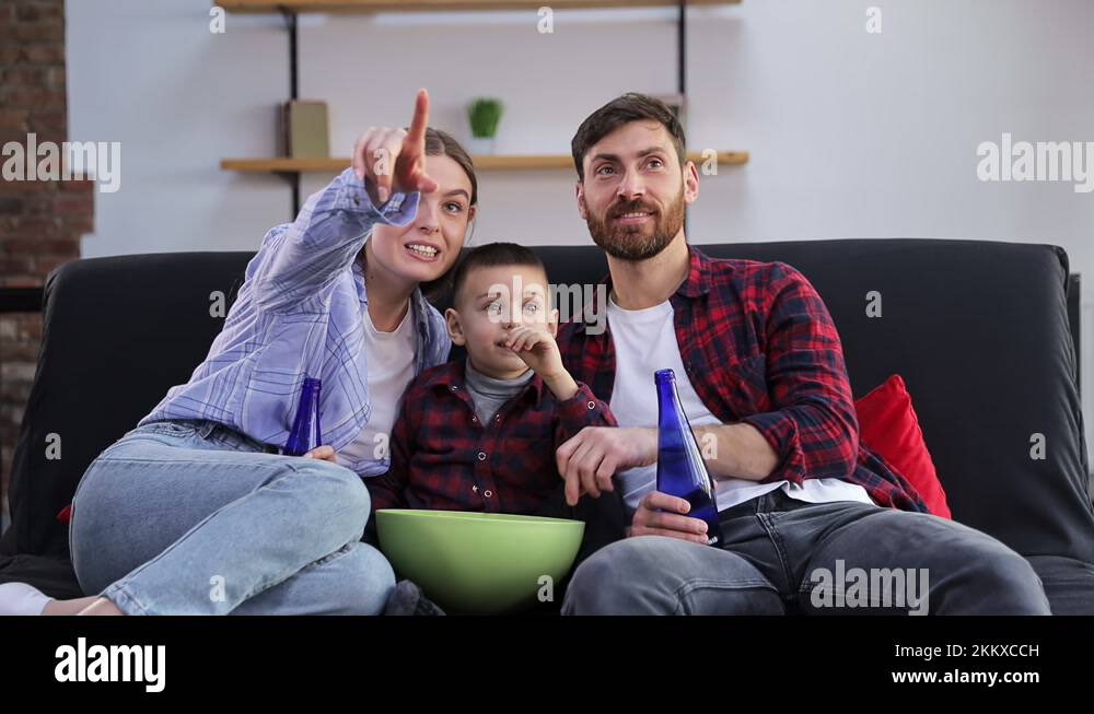 Beautiful young parents, their son is watching TV, laughing, showing ...
