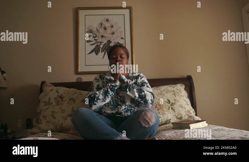 Black woman sitting in bed with legs crossed praying with joy and hope ...