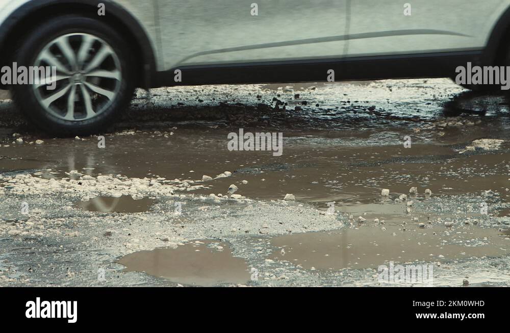 Close-up car driving on a bad road with cracks, muddy puddles and pits ...