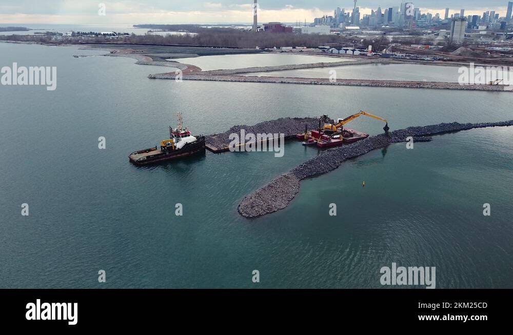 Excavator on barge building breakwater from rocks with Toronto skyline ...