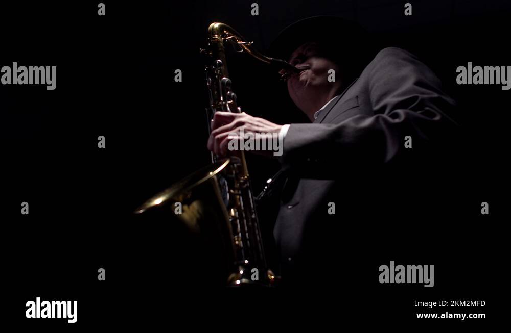 Man saxophonist in hat and suit plays the saxophone in a dark studio ...