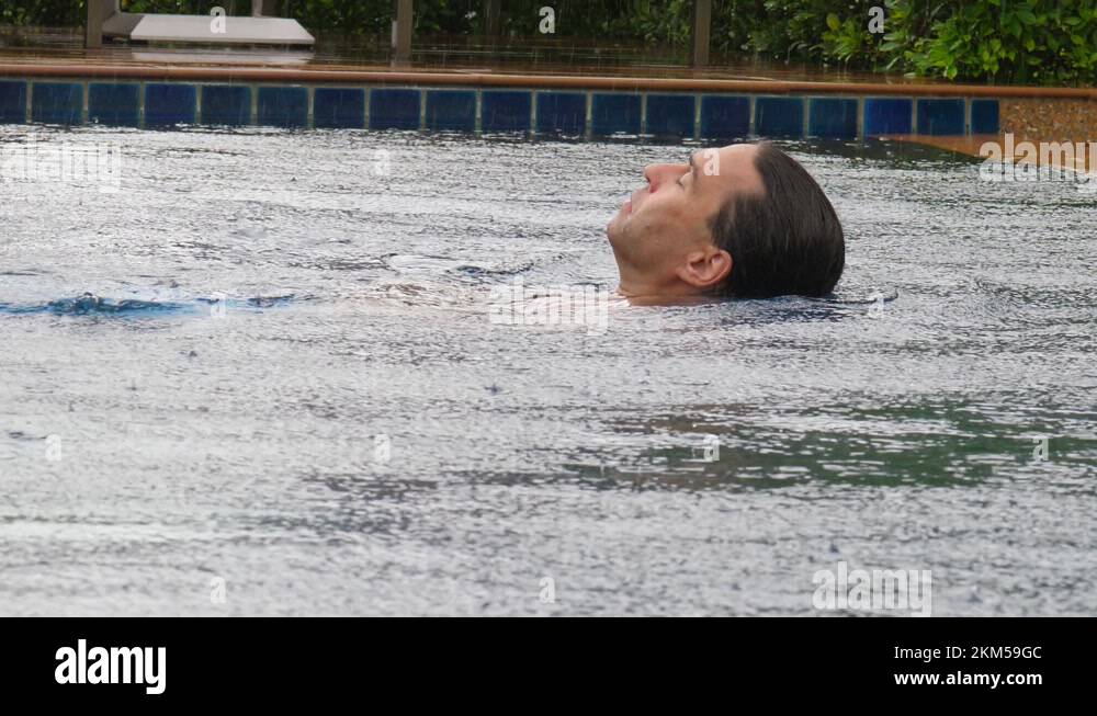 Relaxed tourist man lazily swimming in a pool during a heavy rain ...