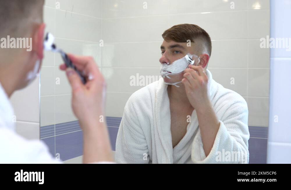 A young man shaves his face hair in front of a mirror. A man in a white ...