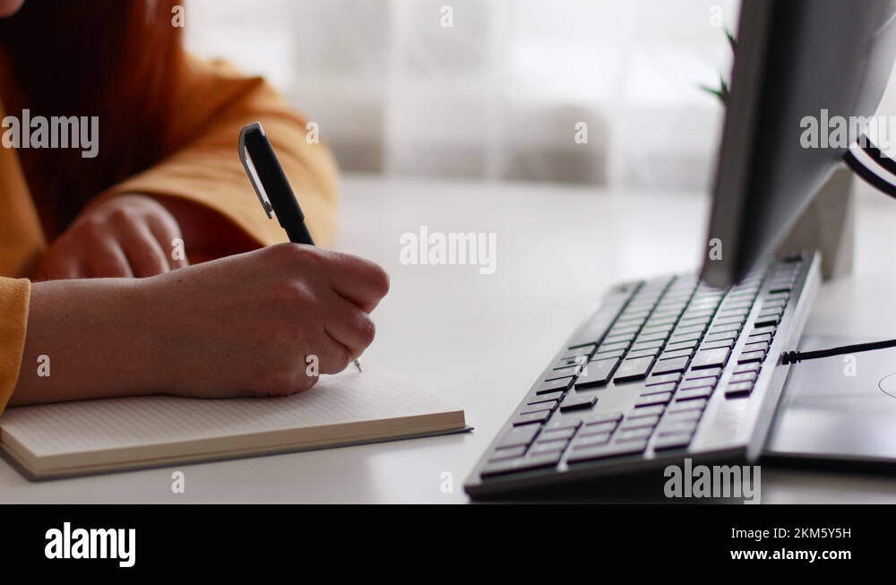 Young girl sit desktop pc hold pen write notepad in home office indoors ...