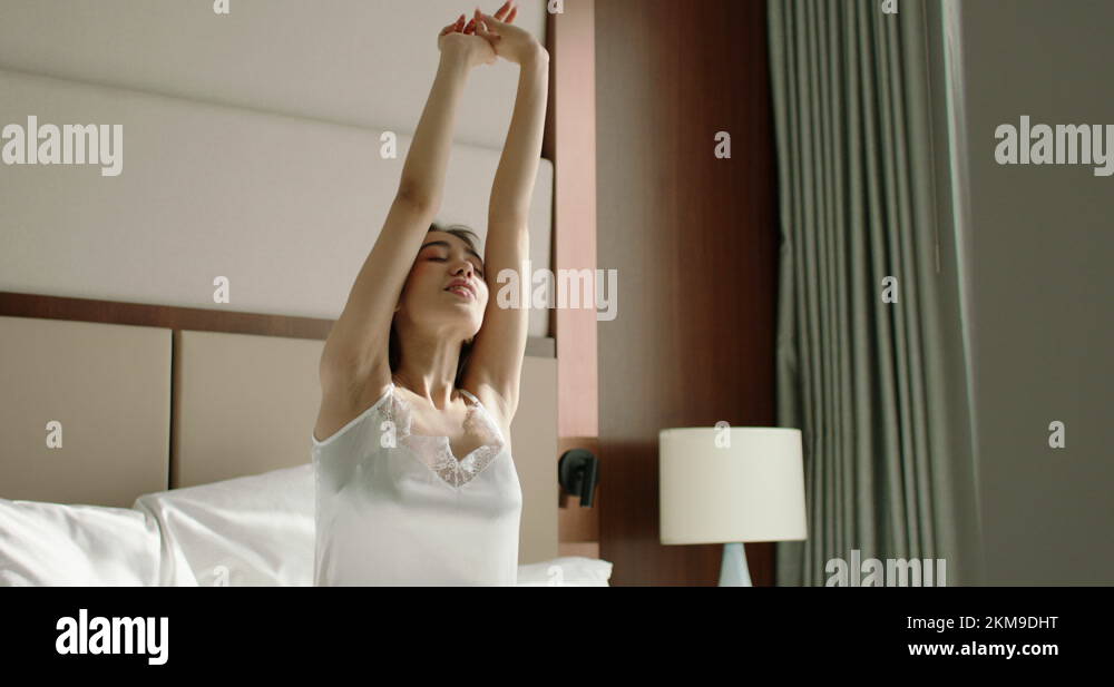 Relaxed asian girl waking up in cozy hotel room, raising her hands up ...