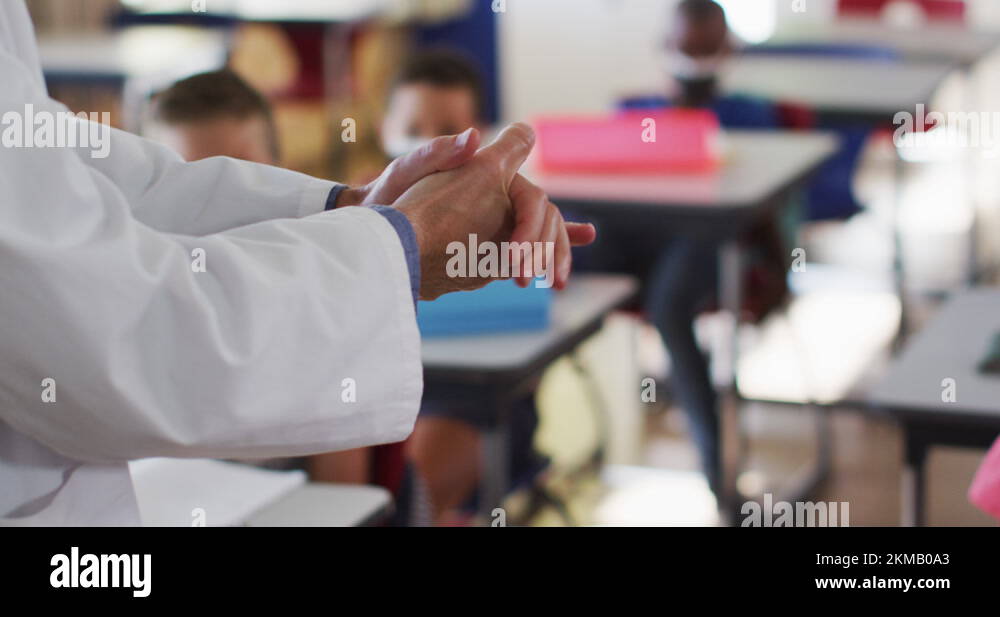 Diverse teacher disinfecting hands in classroom, with schoolchildren ...