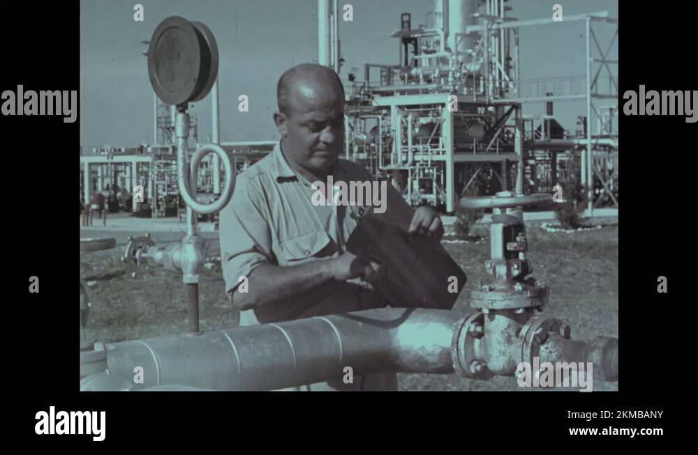 1960s: Man looks at pipe at oil refinery, writes on clipboard. View of ...