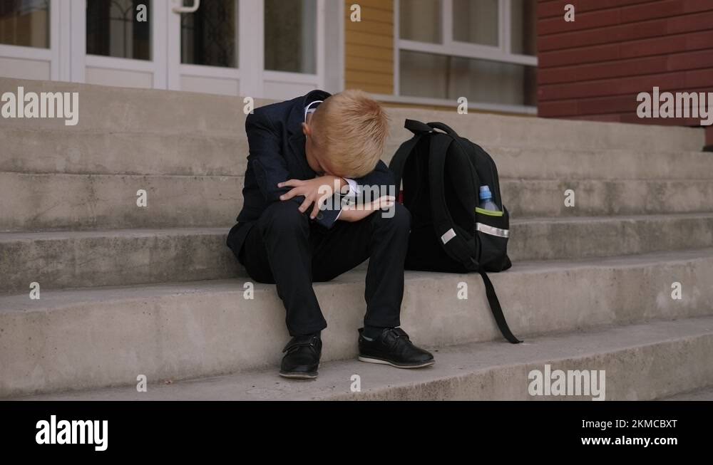 A sad boy cries while sitting on the school stairs, his backpack is ...