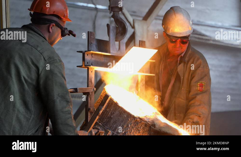 Two men sloping a big metallic barrel with liquid orange iron flowing ...