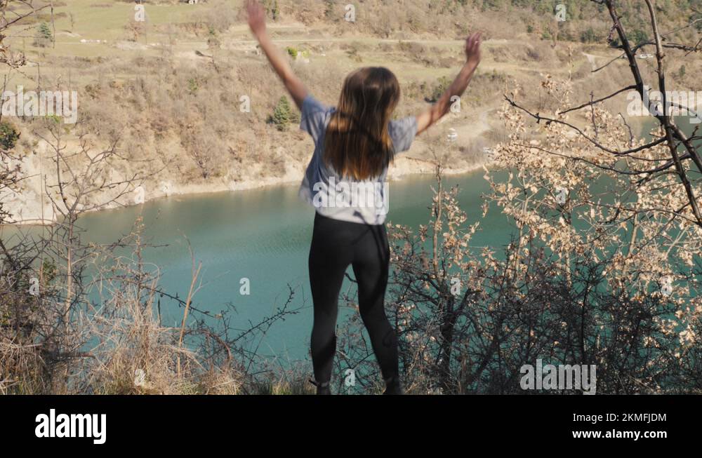 Young woman stands with raised arms up on the nature lake sunset ...