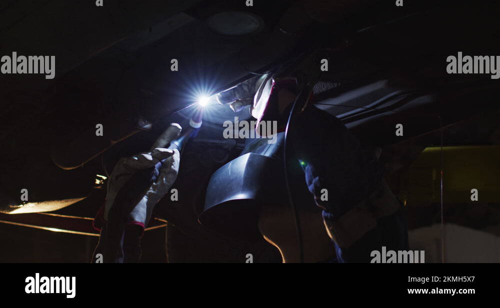 Female mechanic wearing welding helmet welding under a car at a car ...