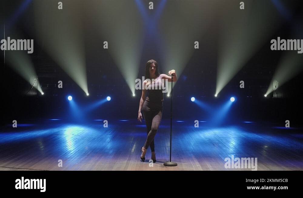 A singer poses on stage in dark under the light of a white and blue ...