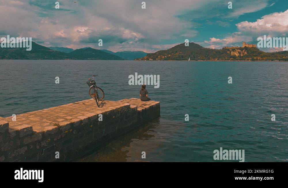Back view of lonely woman with bicycle behind sitting on pier edge on ...