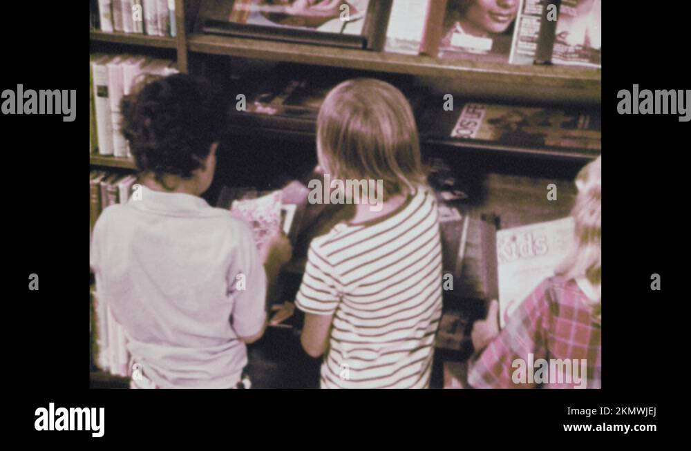 1970s: Several kids look at magazines on shelf in library. Posters on ...