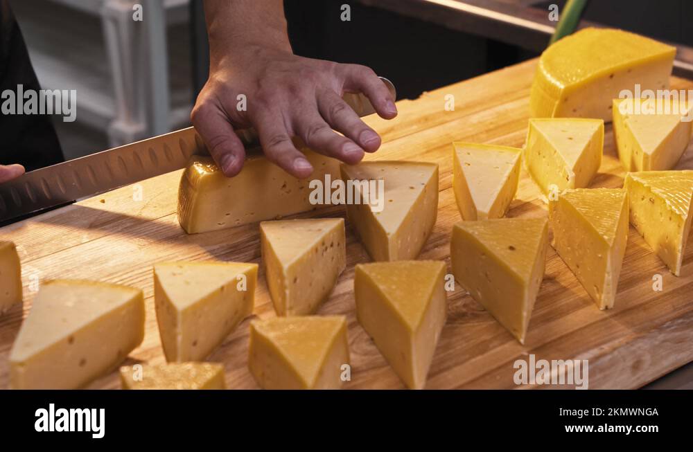 A man in the shop cuts a cheese with a big knife in triangular similar ...