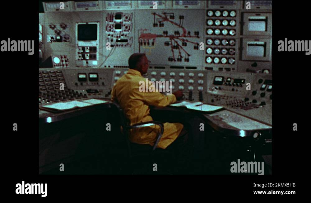 1960s: Man sits at console control panel of cyclotron. Man adjusts ...