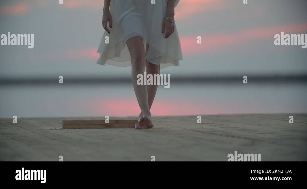 Blonde woman in white dress is walking on the pier of the lake and ...