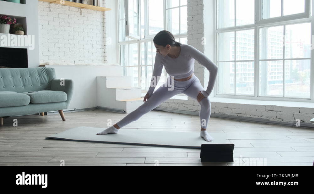 Athletic young woman stretches legs and arms at home with large windows ...