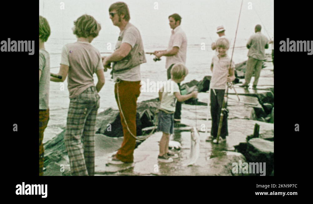 1970s: Waves crash on jetty. Men stand and fish. Boys play with fish ...