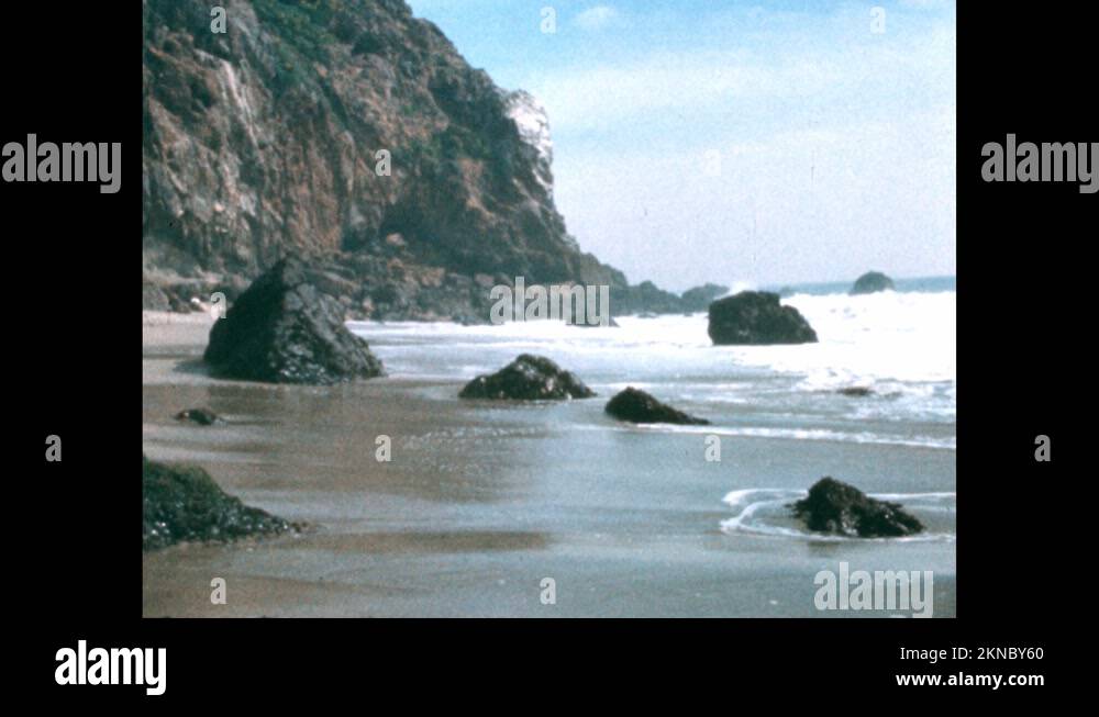 1960s: Rock on string hangs in place. Waves wash across rocks on beach ...