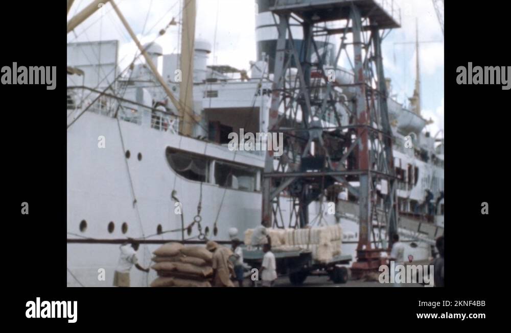 1960s: Men and cranes load cargo onto ship at docks. Man sprays ...