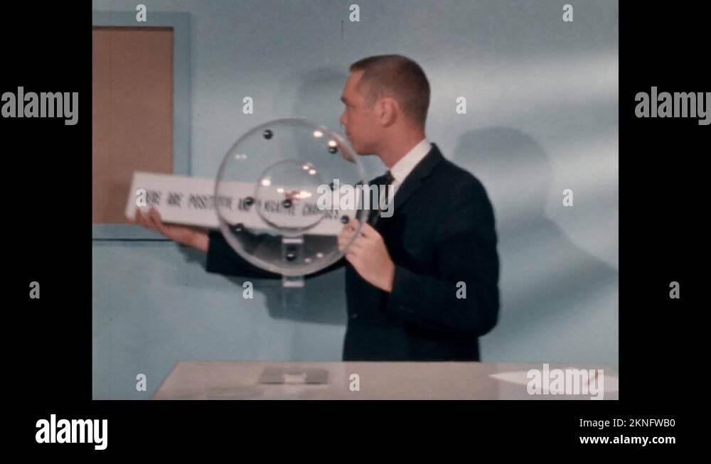 1960s: Man standing with electron model, talking. Man holding long text ...