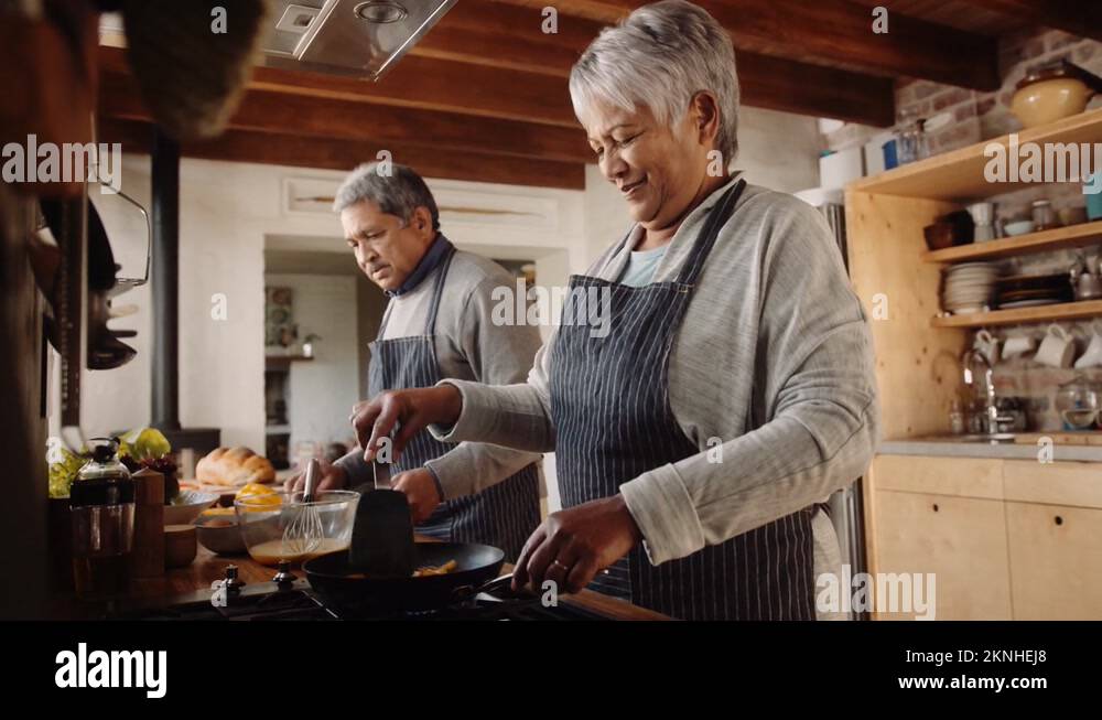 Multi-cultural elderly woman cooking in kitchen, helped by husband ...