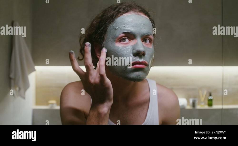 Young man smears a clay mask on his face, looking into camera as in a ...