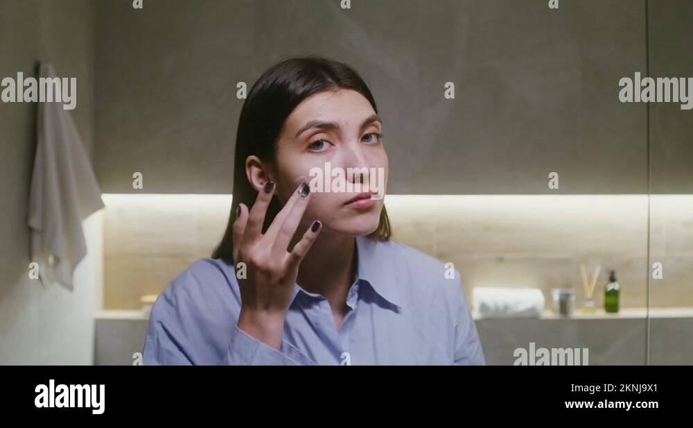 Young woman smears moisturizer on her face, looking into camera as in a ...