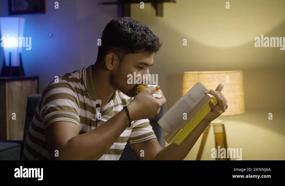 young indian student eating unhealthy food while reading book at home ...