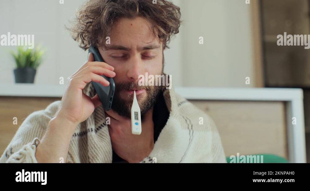 Sick young man with termometr in mouth talking on the phone while ...