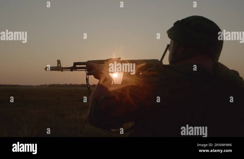 Uniformed soldier sniper with assault rifle aiming at enemy at sunset ...