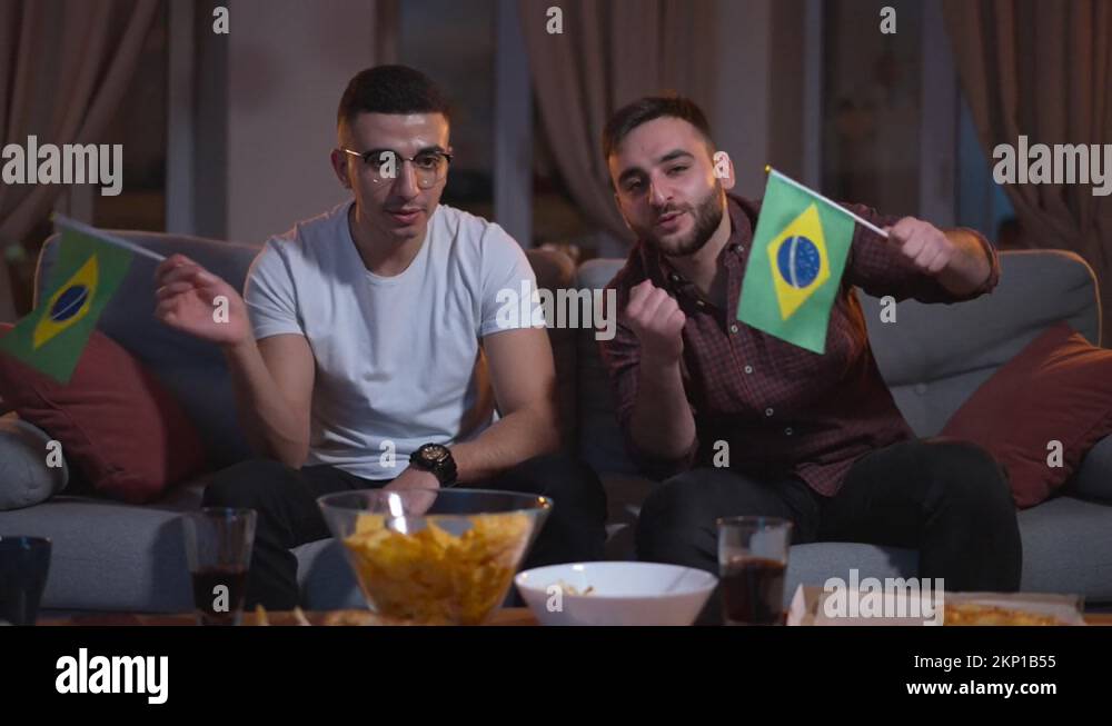 Two men watching tv match and waving brazil flags, sitting on couch in ...
