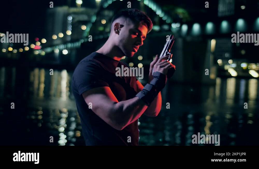 A young gangster holding a gun with city lights in background at night ...