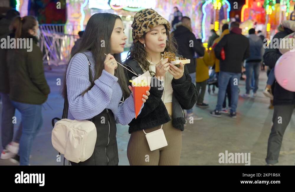 Two young women talking while eating snacks at night Valencia funfair ...