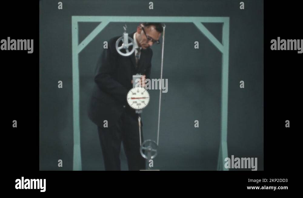 1960s: Man holds a spring scale attached to a rope and pulley system ...