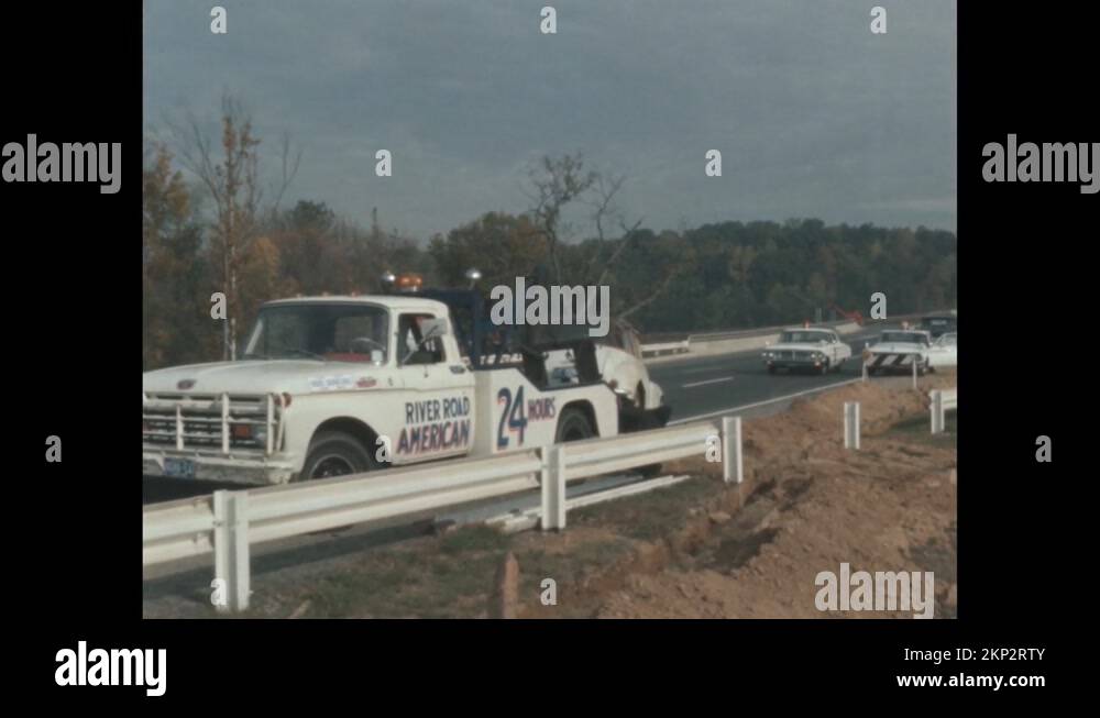 1960s: Person operating tow truck on roadside. Person lifting car onto ...